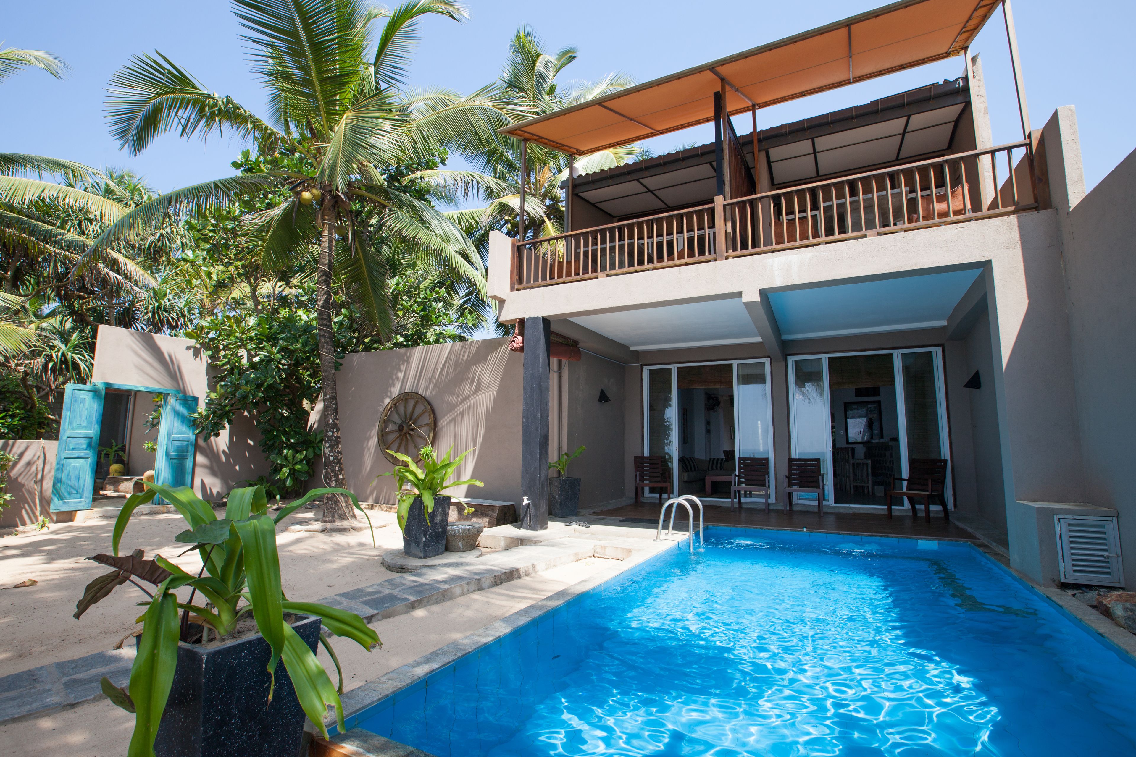 Uma área de piscina moderna com um edifício de dois andares, varanda coberta e palmeiras ao redor.