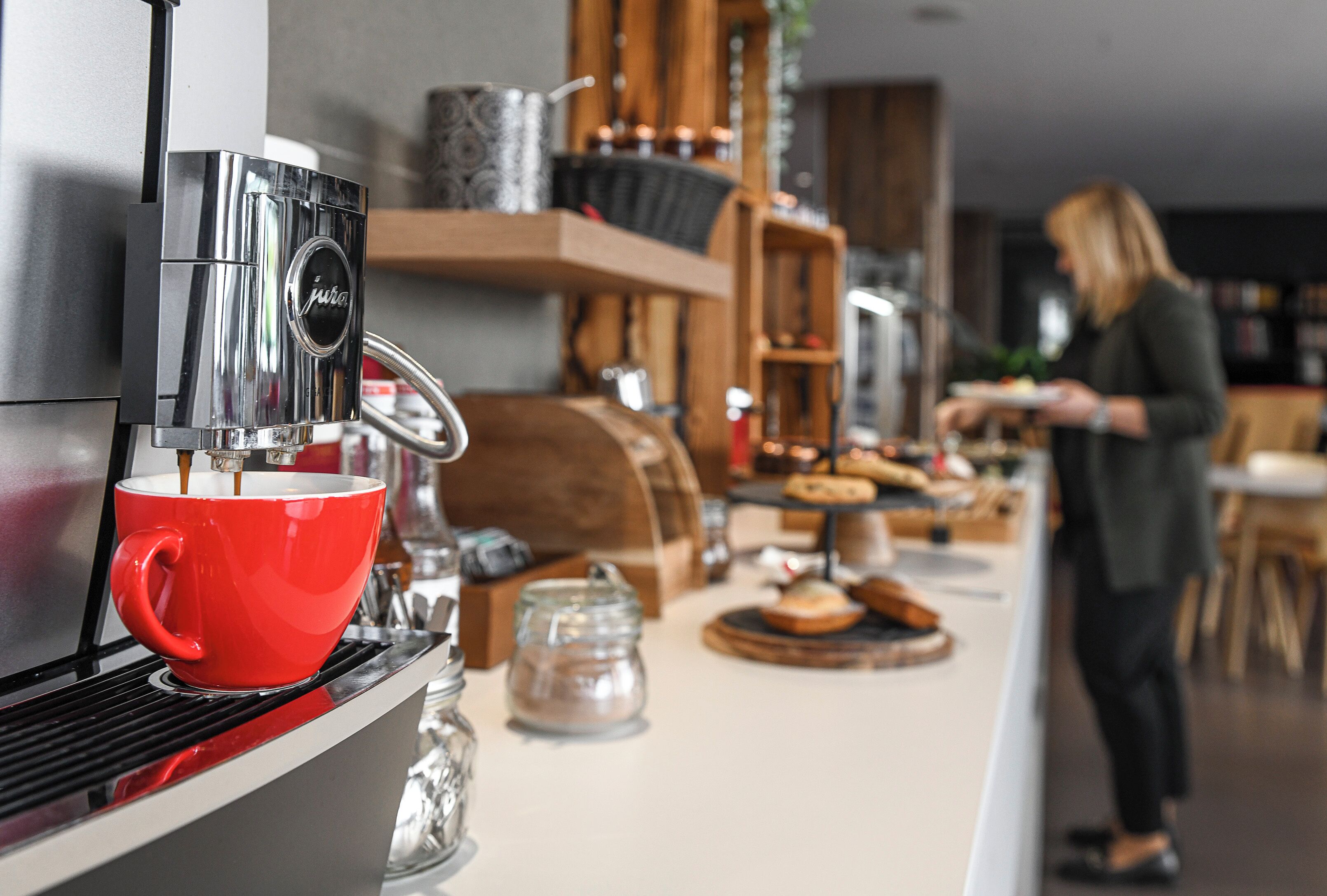 Máquina de café a servir expresso numa chávena vermelha, com um balcão a exibir pastelaria e uma mulher ao fundo.