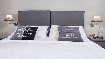 Cama bem arrumada com duas almofadas, cada uma com uma citação inspiradora, e duas luminárias fixadas nas paredes, uma de cada lado.