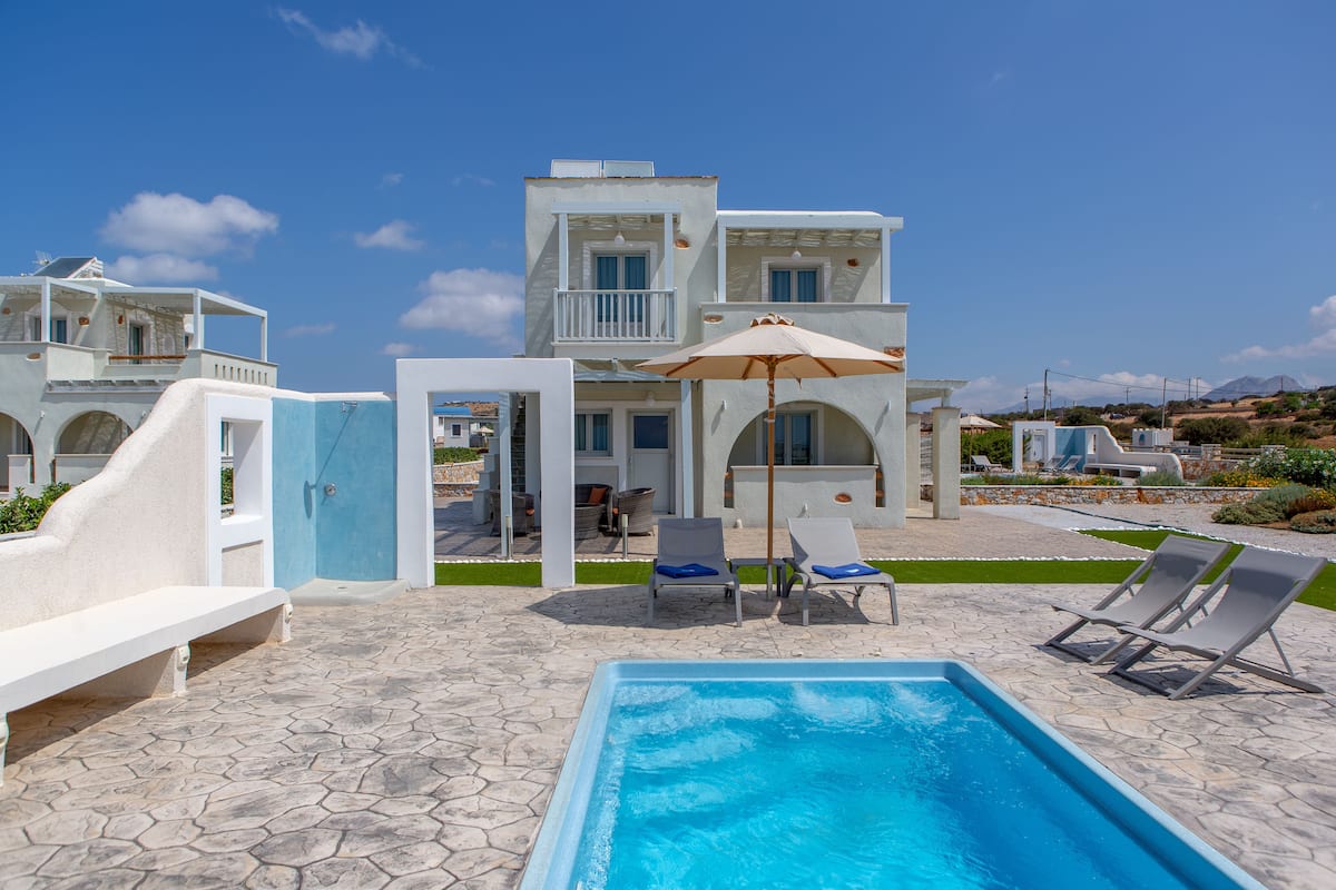 Uma área de piscina com espreguiçadeiras e um banco, um edifício com varanda, e um céu azul e límpido.