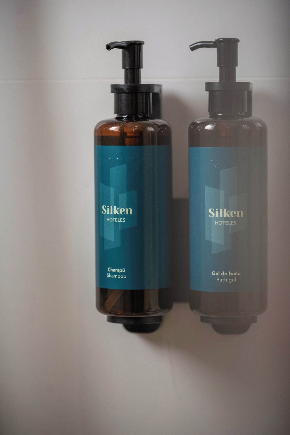 Dois frascos de produtos de higiene do hotel Silken, um com a etiqueta 'Champú Shampoo' e outro 'Gel de banho Bath gel', fixados na parede.