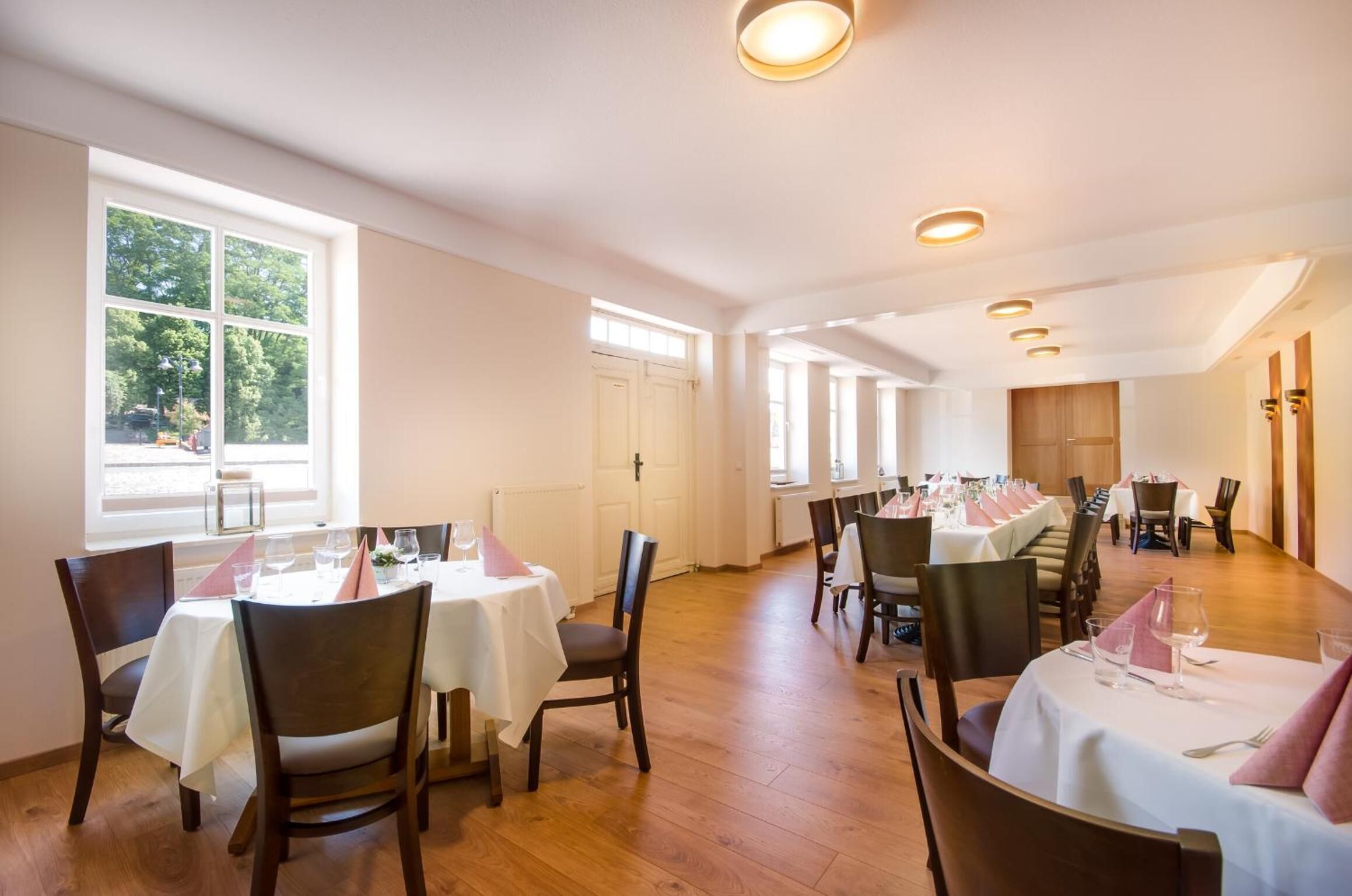 Uma sala de jantar com mesas redondas, cadeiras e vista para o exterior através de amplas janelas.