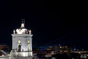 Uma torre do relógio branca com cúpula e cruz no topo, rodeada por edifícios.
