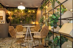 Uma área de jantar acolhedora, com cadeiras de vime e uma mesa redonda, rodeada por plantas verdes exuberantes e um chão com padrão geométrico.
