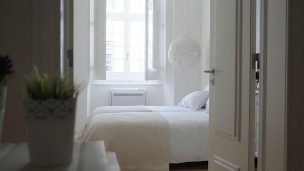 Quarto minimalista com uma janela grande, uma cama com roupa de cama branca e uma planta em vaso sobre uma mesa de cabeceira.