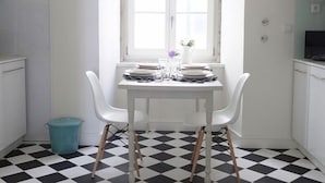 Uma cozinha pequena com uma mesa branca posta para dois, uma janela acima da mesa e piso xadrez.