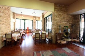 Uma sala de jantar ampla, com mesas e cadeiras de madeira, paredes de pedra e grandes janelas que permitem a entrada de luz natural.