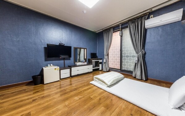 Quarto de hotel com piso de madeira, uma cama, escrivaninha com computador, televisão na parede e janela com cortinas.