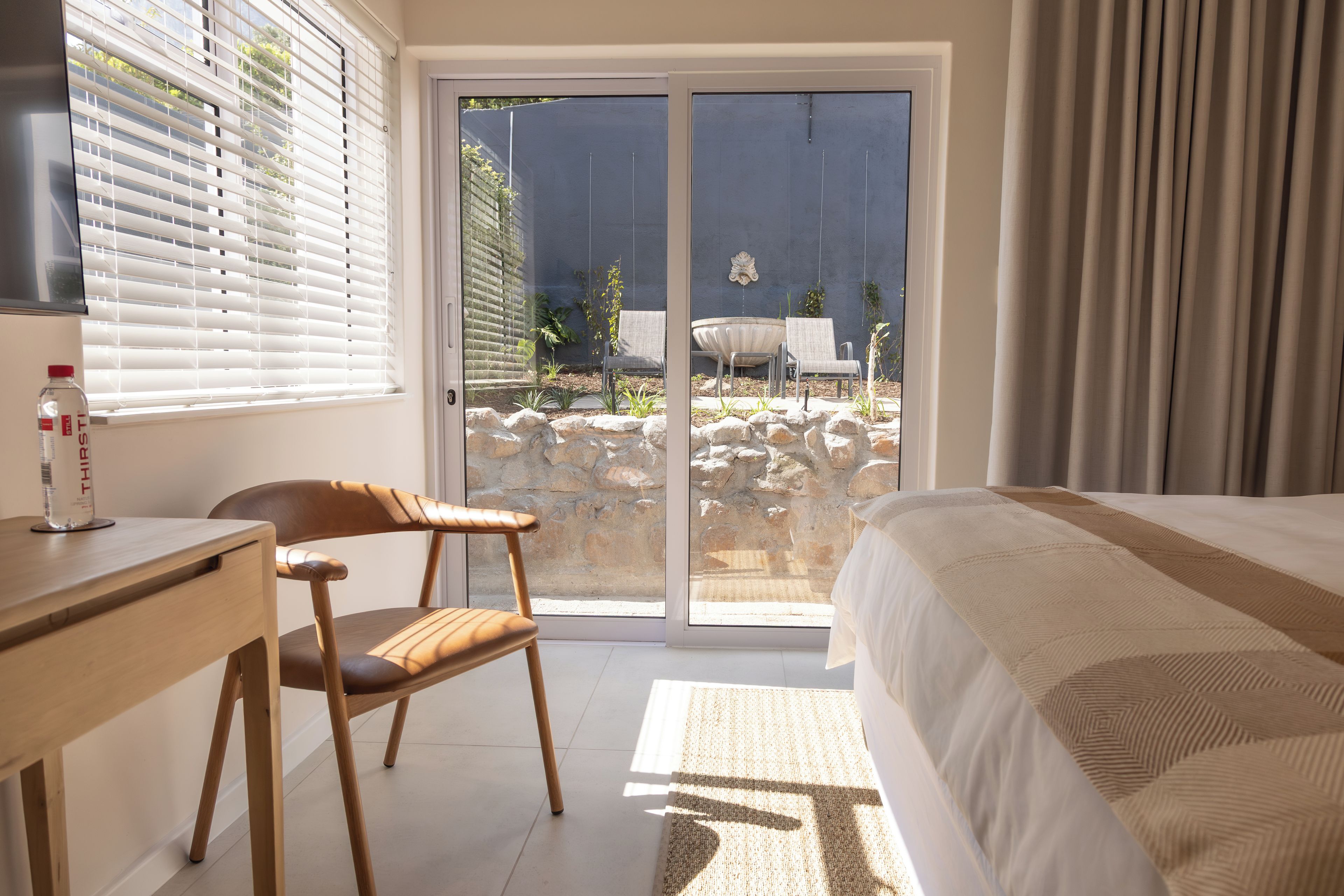 Quarto com cama, escrivaninha, cadeira e vista para área externa com piscina e plantas.