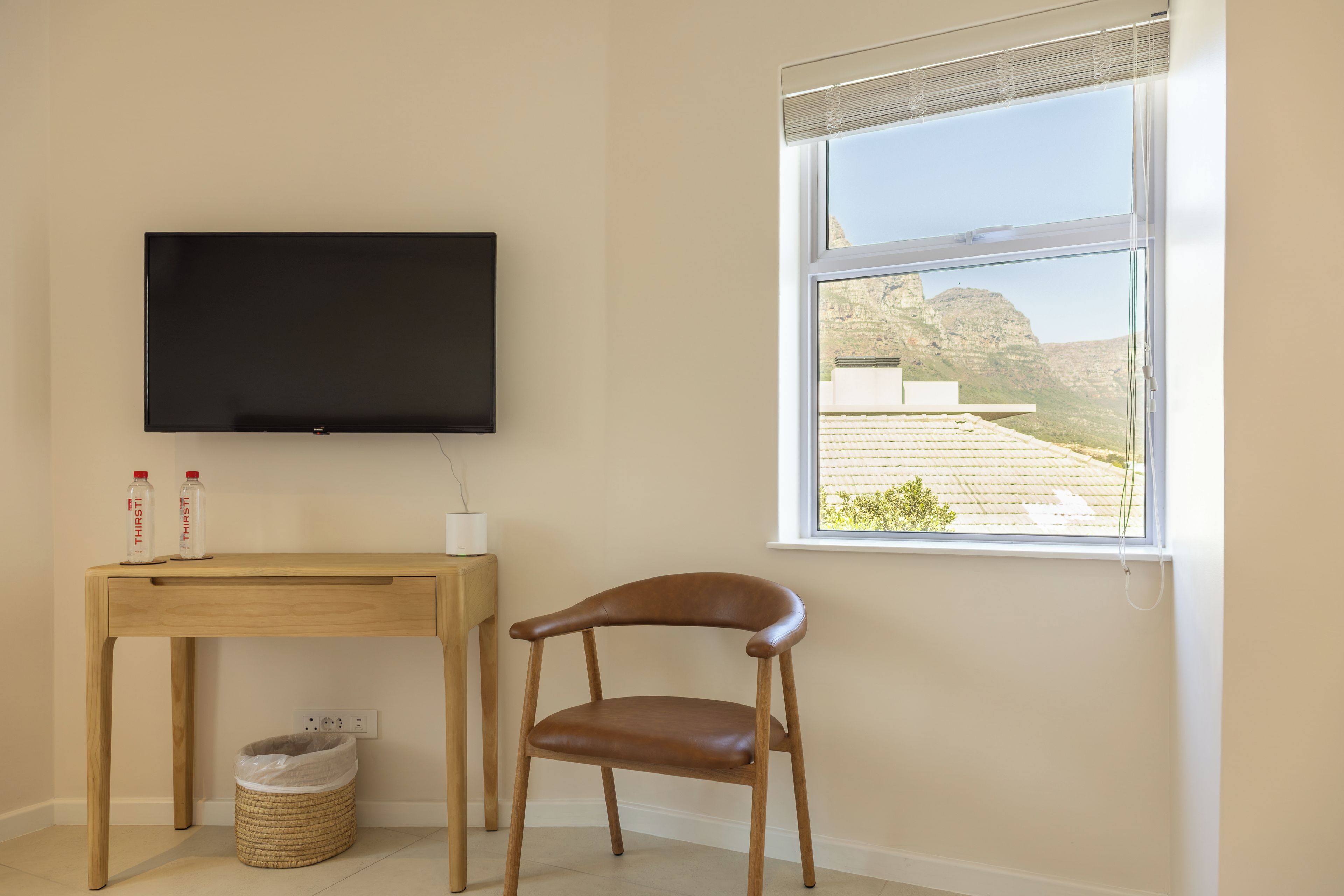 Quarto com escrivaninha de madeira, poltrona de couro marrom, televisão fixada na parede e janela com vista para uma paisagem montanhosa.