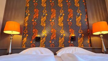 Quarto de hotel com papel de parede estampado, duas luminárias de cabeceira e uma cama com lençóis brancos.