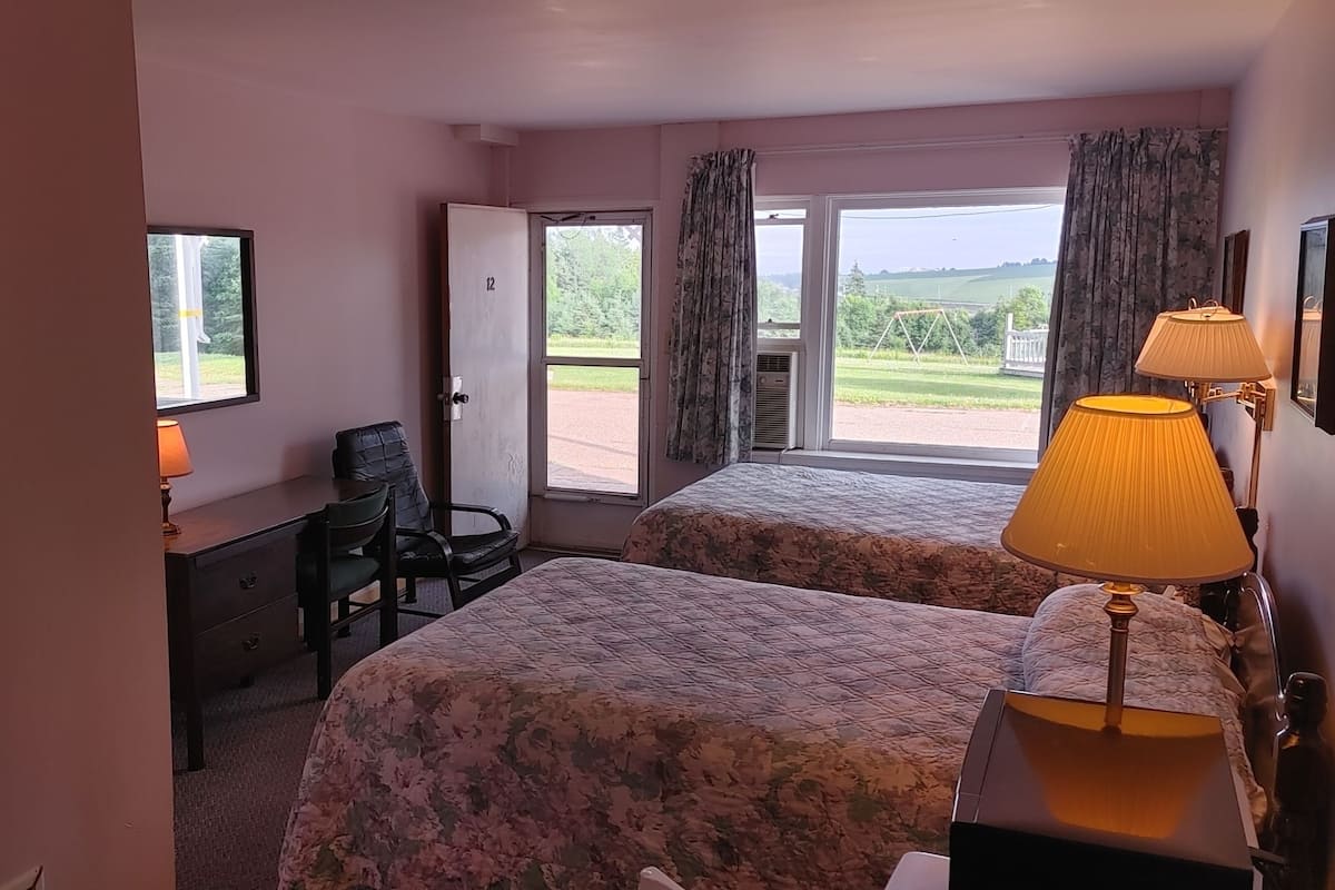 Quarto de hotel com duas camas, uma escrivaninha, uma cadeira e uma janela com cortinas.