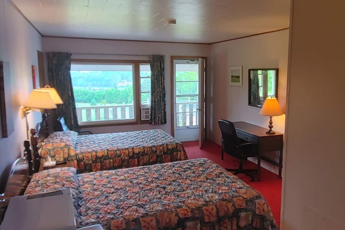 Quarto de hotel com duas camas, uma escrivaninha e uma cadeira. Há uma janela com vista para vegetação lá fora.