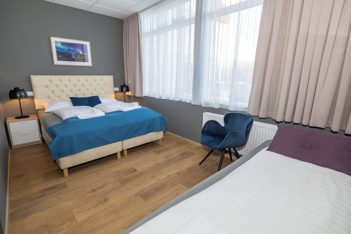 Quarto de hotel com uma cama grande, uma cadeira, mesa de cabeceira, abajur e janela com cortinas.