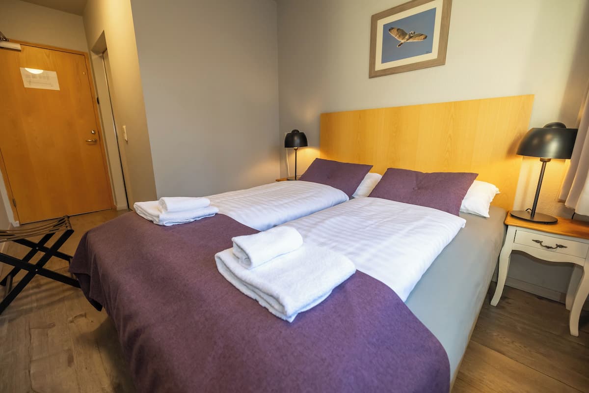 Quarto de hotel com duas camas, cabeceira de madeira, mesa de cabeceira com abajur e quadro na parede.