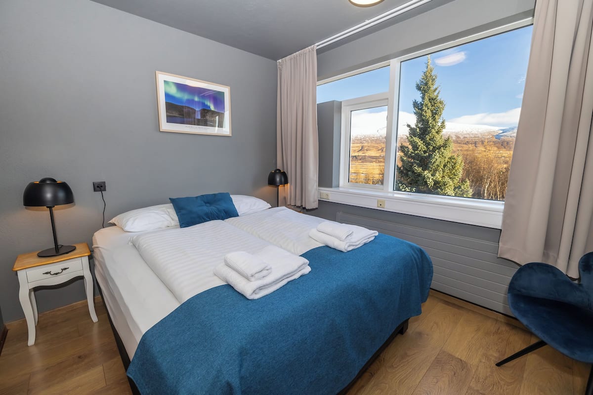 Cama bem arrumada com roupa de cama azul e branca, mesa de cabeceira com abajur, e vista para uma paisagem nevada através de uma janela.