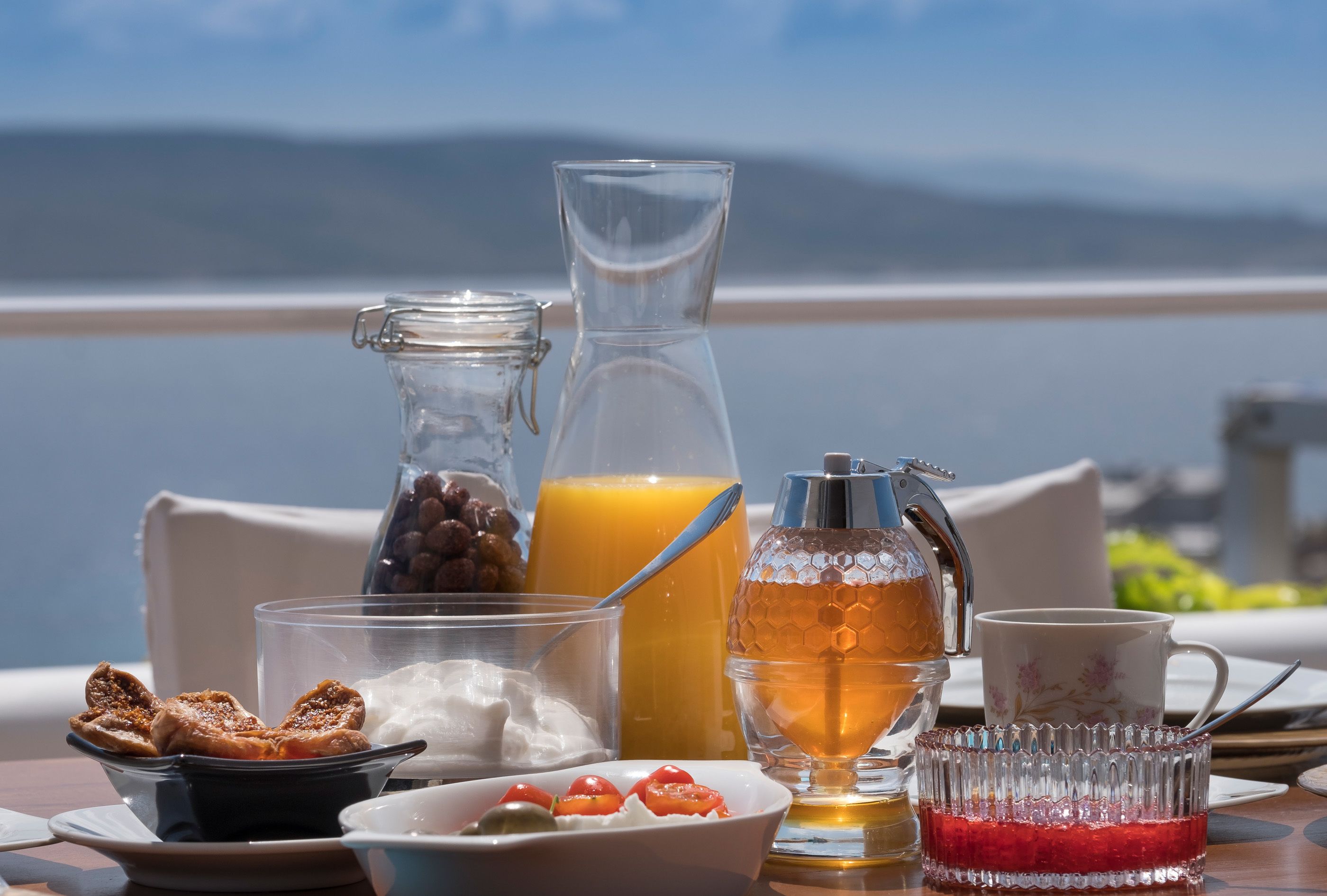 Mesa de café da manhã com diversos alimentos e bebidas, um jarro de vidro e vista clara do oceano ao fundo.