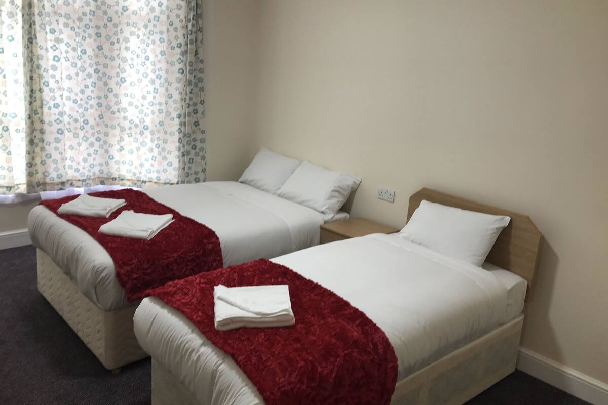 Quarto de hotel com duas camas, cada uma com roupa de cama branca e um cobertor vermelho. Há uma janela com cortinas florais e uma pequena mesa de cabeceira de madeira com um abajur entre as camas.