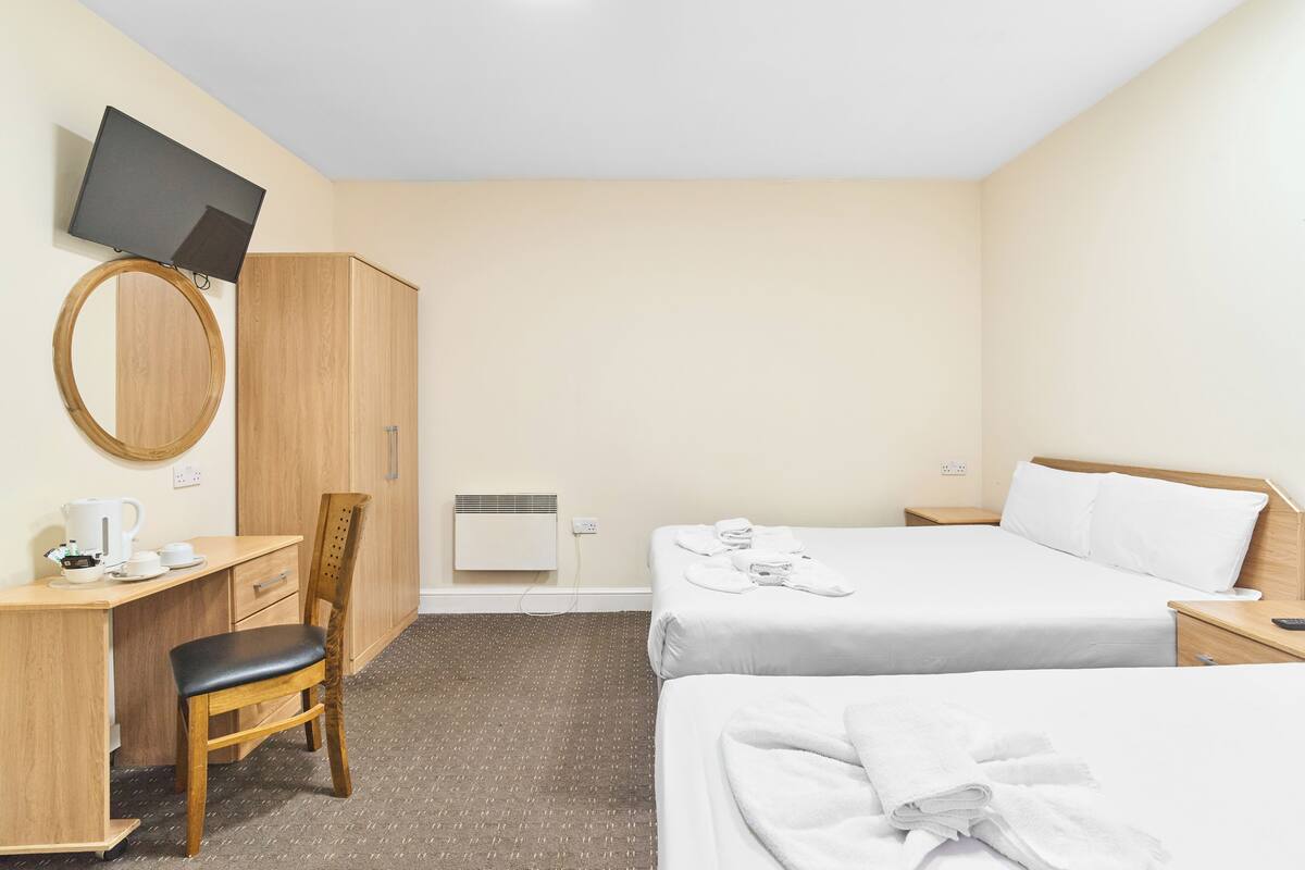Quarto de hotel com duas camas, uma escrivaninha com cadeira, uma TV fixada na parede e um espelho redondo.