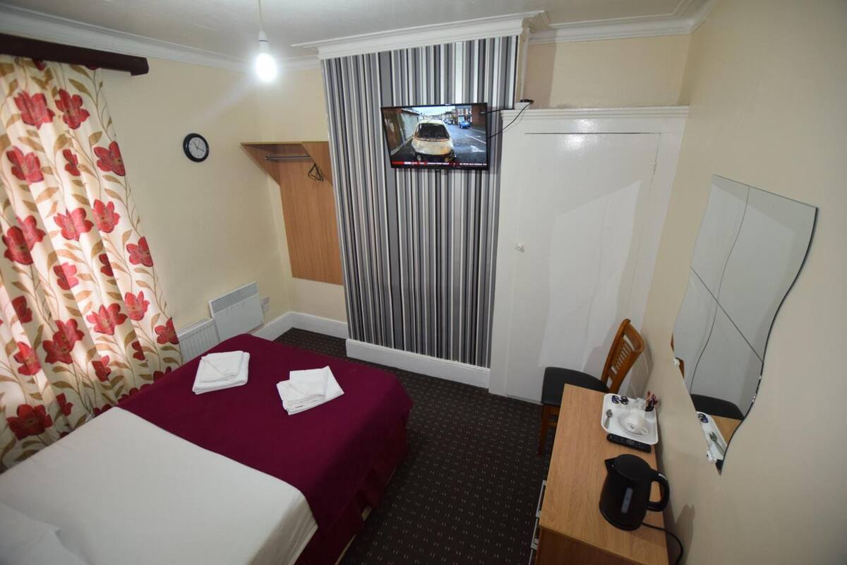 Quarto de hotel com cama, secretária com chaleira, televisão na parede e janela com cortinas.