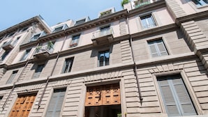 Um edifício de vários andares com estilo arquitetônico clássico, apresentando uma entrada central com porta de madeira, diversas janelas com persianas e varandas com plantas.