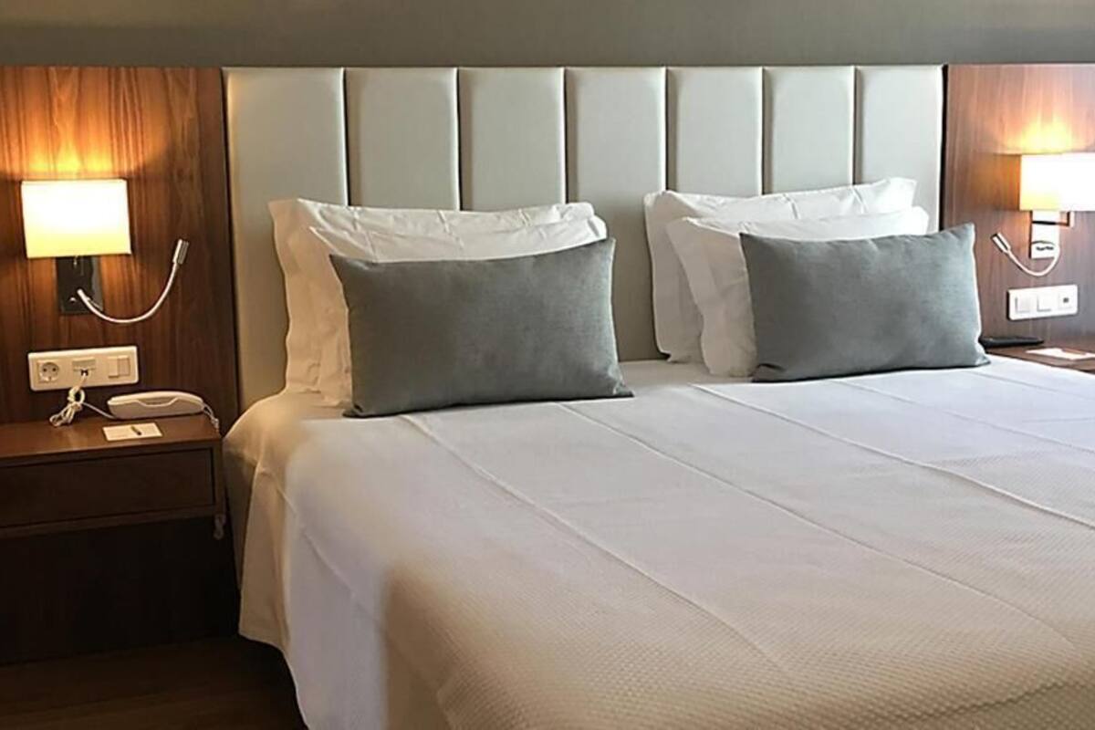 Quarto de hotel com uma cama grande, duas mesas de cabeceira e luminárias fixadas na parede.