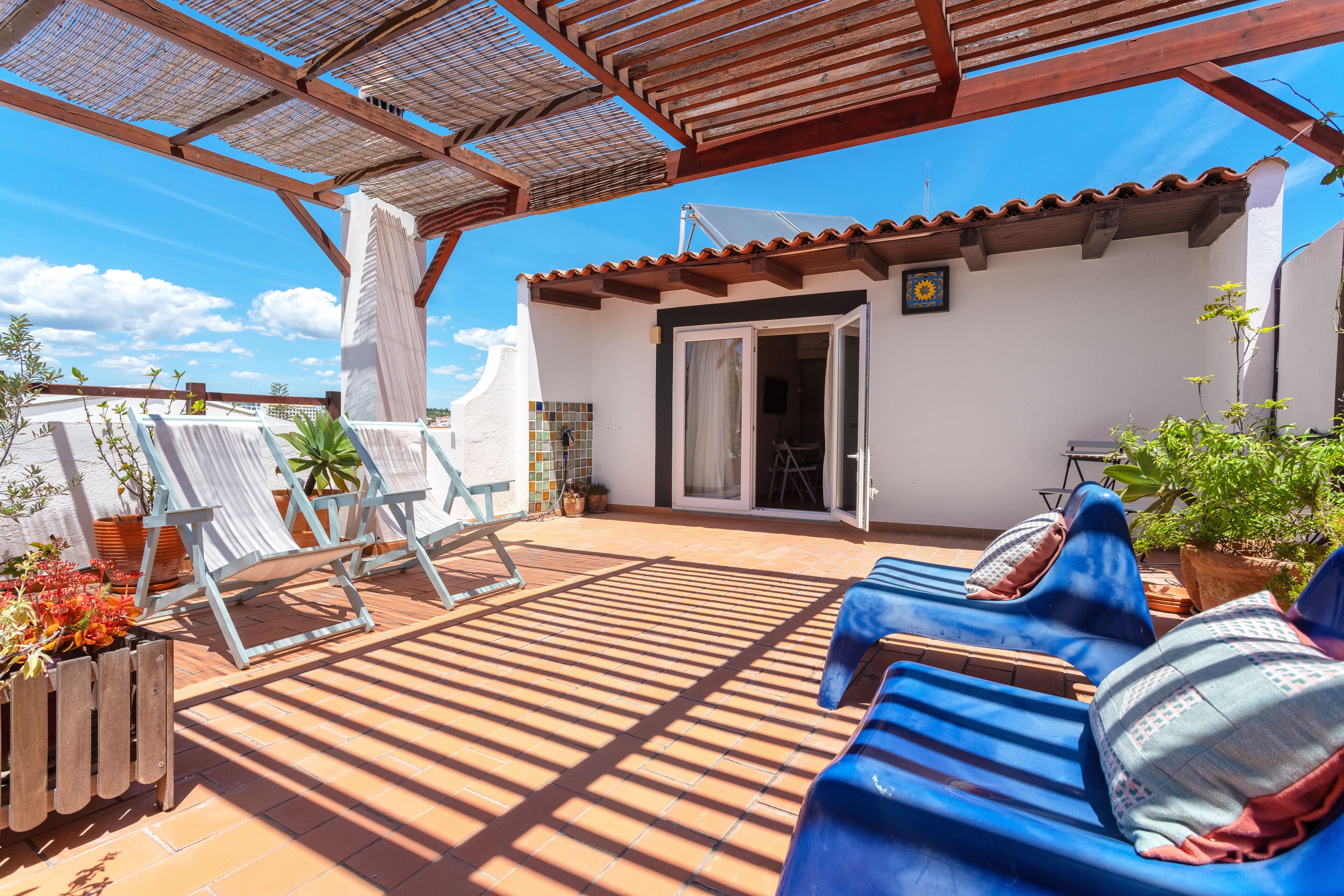 Uma área externa coberta com cadeiras de descanso azuis, uma pérgola de madeira e piso de azulejos.