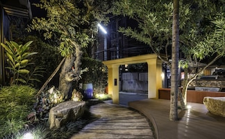 Área externa bem iluminada com um caminho de madeira que leva a uma entrada coberta, cercada por vegetação exuberante e pedras grandes.