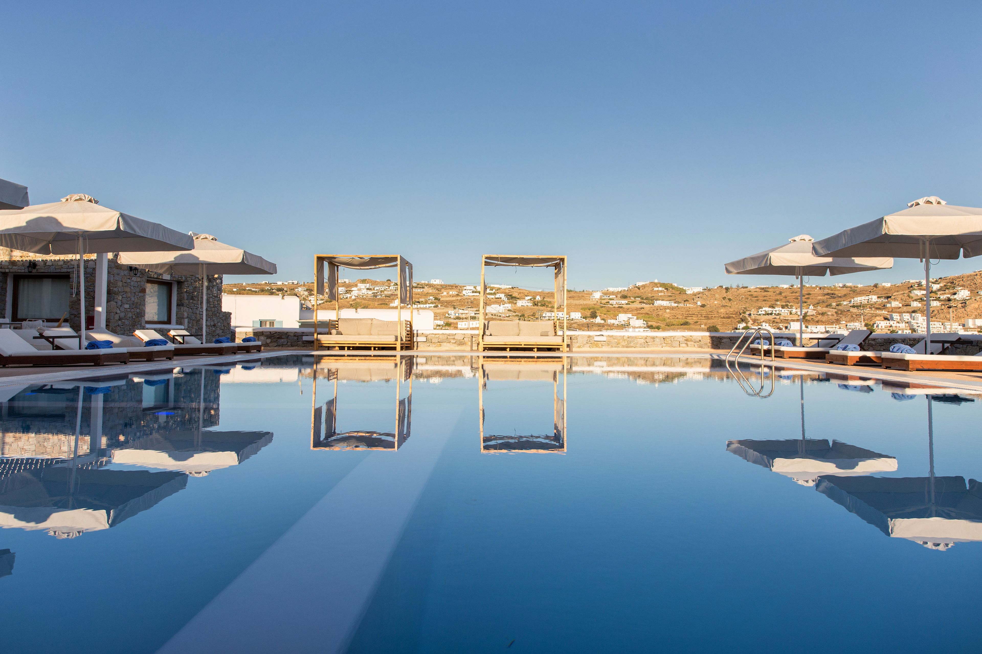 Piscina límpida com espreguiçadeiras e guarda-sóis, rodeada por edifícios de pedra e uma paisagem montanhosa.