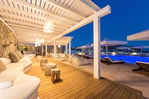 Um deck de madeira com cadeiras de descanso e área de piscina, apresentando uma parede de pedra e mobiliário branco.