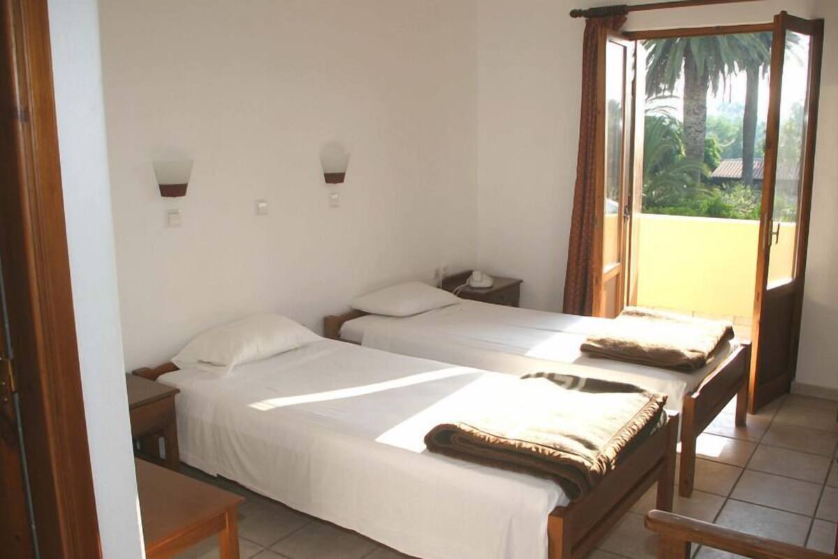 Quarto com duas camas, um banco de madeira, uma mesinha de cabeceira e uma porta de correr para uma varanda.