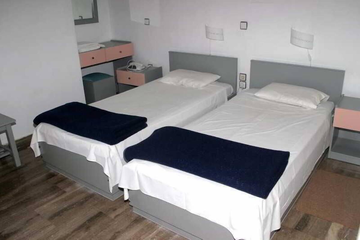 Uma cama de casal com colcha branca e azul, uma mesa de cabeceira, um espelho e uma cômoda pequena com tampo rosa.