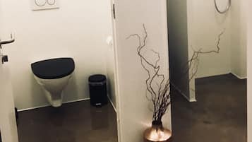 Casa de banho com sanita, lavatório e chuveiro. Há estantes com objetos decorativos e um cesto de lixo.