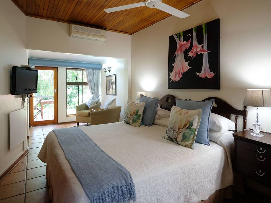 Quarto com cama grande, ventilador de teto, televisão, mesa de cabeceira, abajur e pintura de flores.