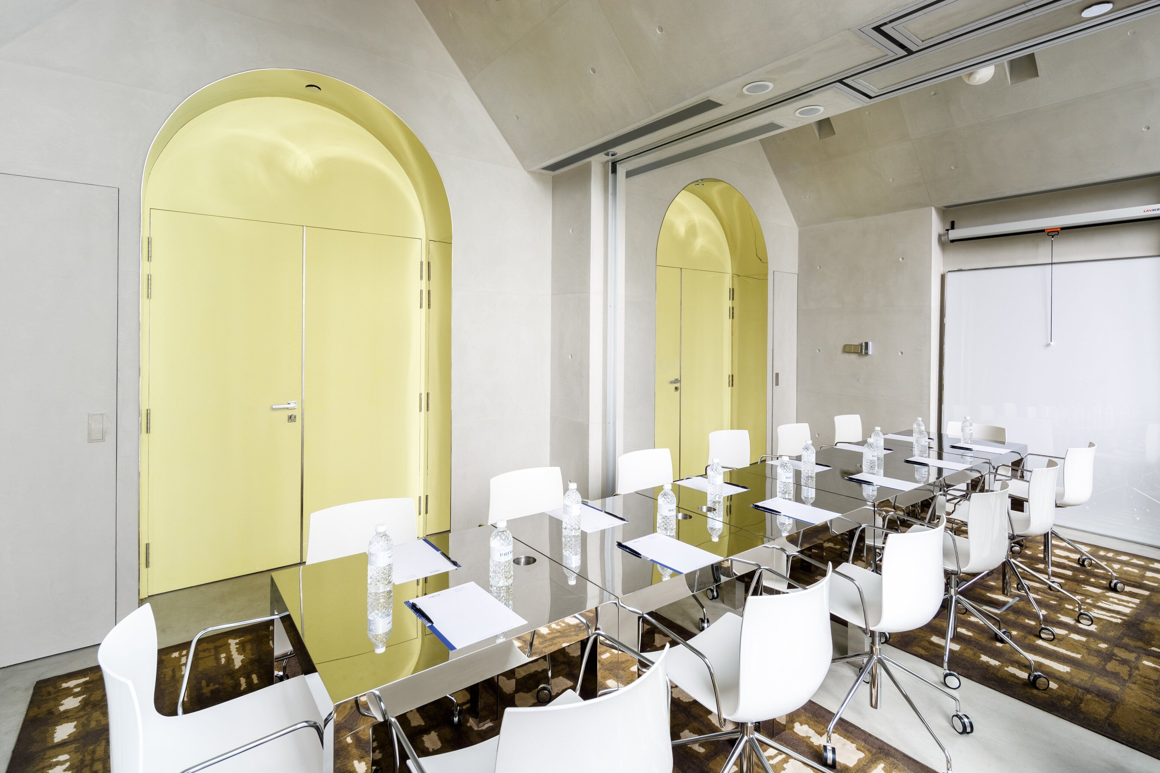 Uma sala de conferência com mesa de vidro, cadeiras brancas, portas amarelas e piso coberto de carpete.