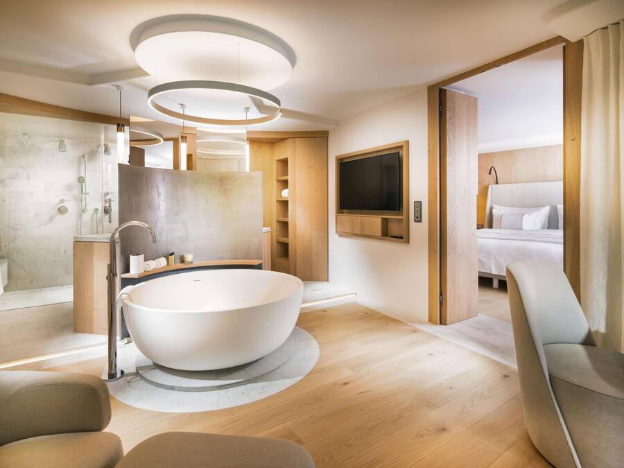 Quarto de hotel moderno com uma banheira grande, uma televisão fixada na parede e uma área de quarto separada com uma cama.