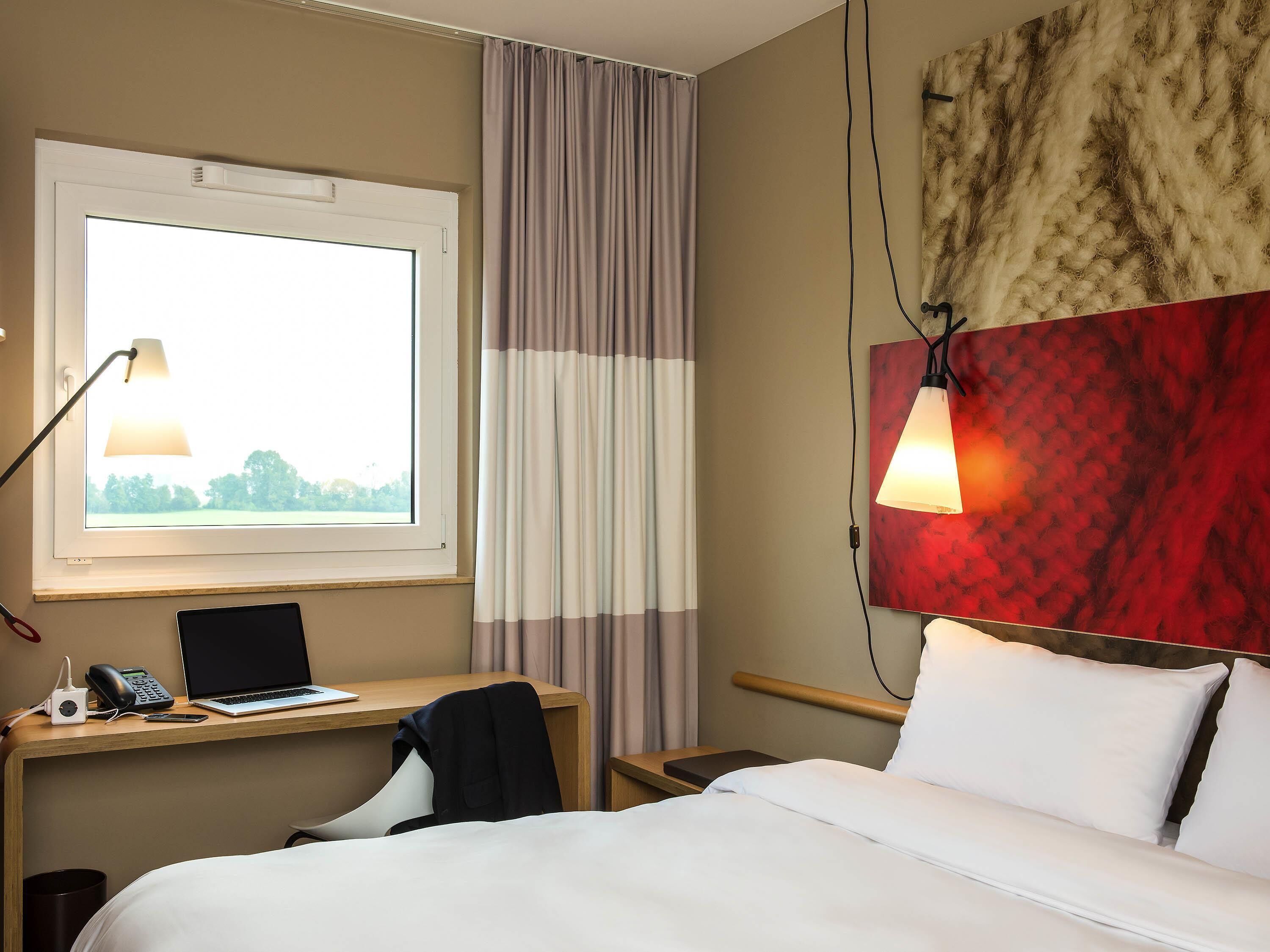 Quarto de hotel com uma cama, uma escrivaninha com um laptop, um telefone e uma luminária.