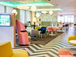 Um hall de hotel moderno com cadeiras coloridas, uma tela grande exibindo um cardápio e um piso estampado.