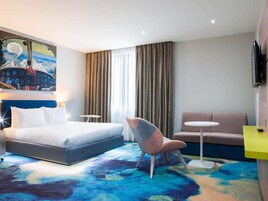 Quarto de hotel moderno com uma cama grande, uma escrivaninha, um sofá e um tapete colorido e abstrato.