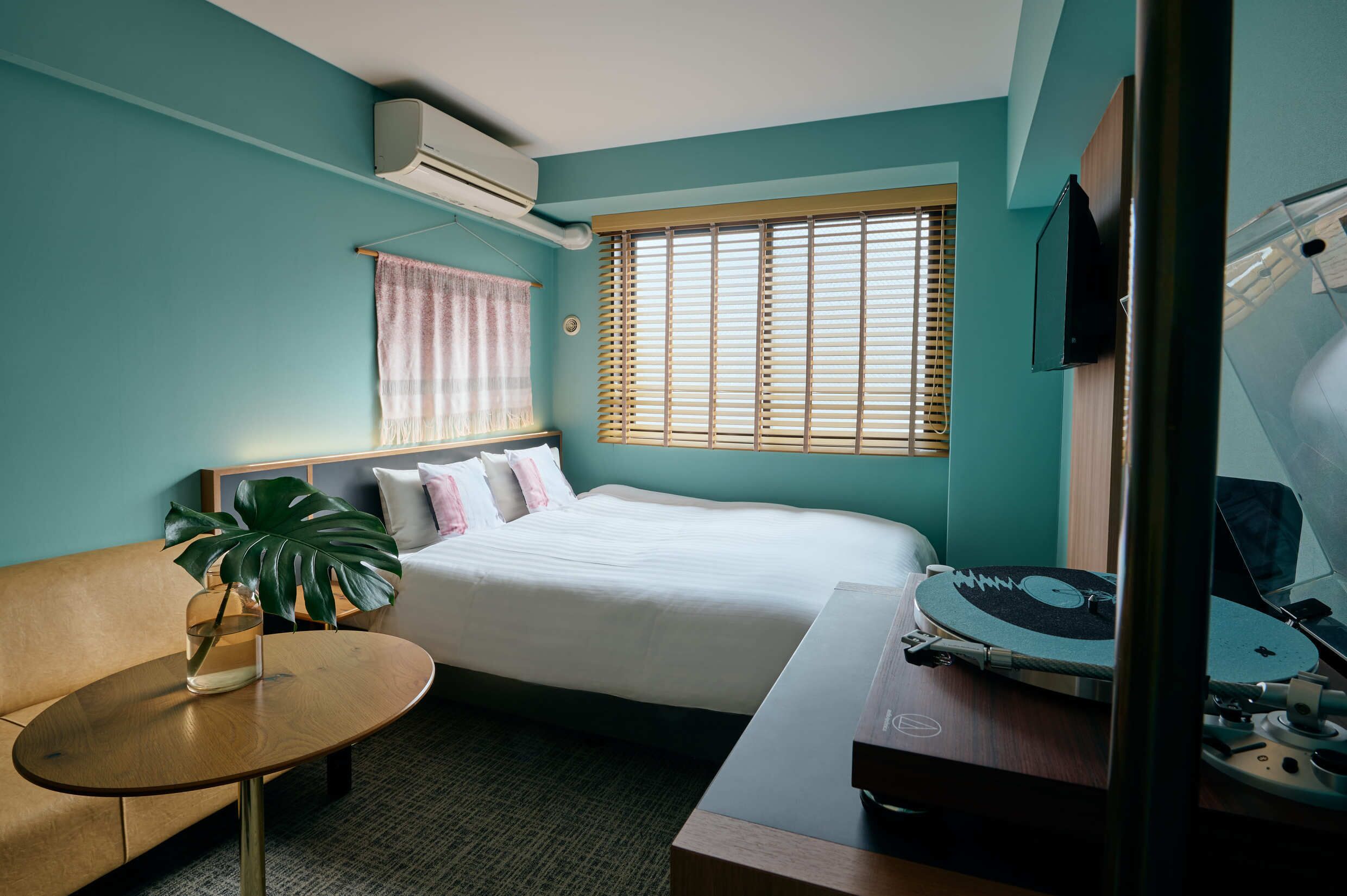 Quarto de hotel com uma cama, uma mesinha, uma planta, uma janela com persianas e uma televisão.