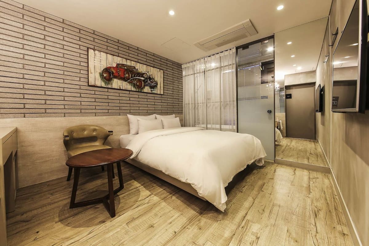 Um quarto com uma cama, uma cadeira, uma mesinha e uma parede com padrão de tijolos, além de uma porta.