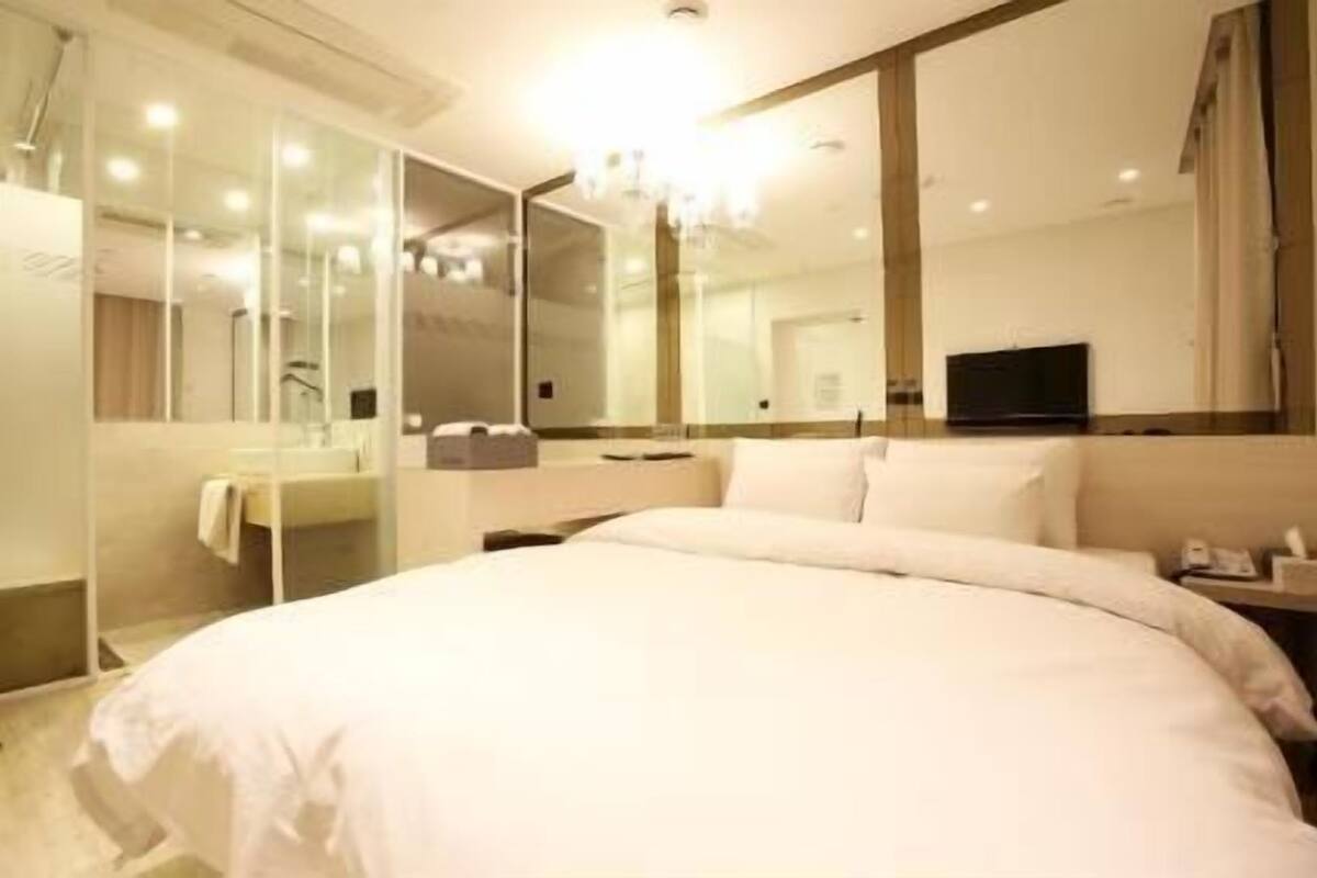 Quarto de hotel com uma cama grande, um espelho, uma televisão e banheiro com chuveiro e pia.