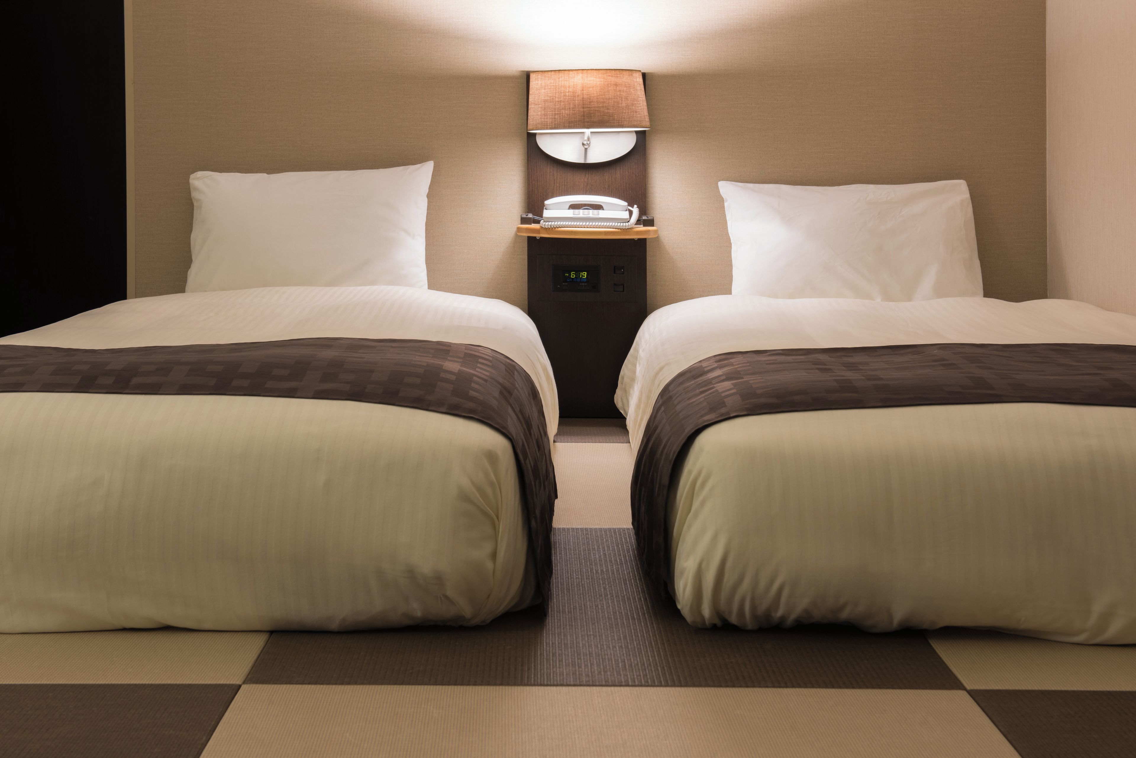 Quarto de hotel com duas camas, uma mesa de cabeceira com abajur e um relógio digital.