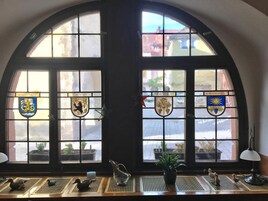Quarto com uma grande janela em arco mostrando quatro brasões, uma pequena mesa com objetos decorativos e uma planta em vaso.
