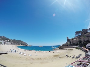 Uma praia com céu claro, uma grande massa de água e uma fortificação histórica numa colina.