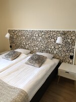 Uma cama de casal com duas almofadas decorativas, uma mesa de cabeceira com abajur e um painel de madeira com padrão circular.