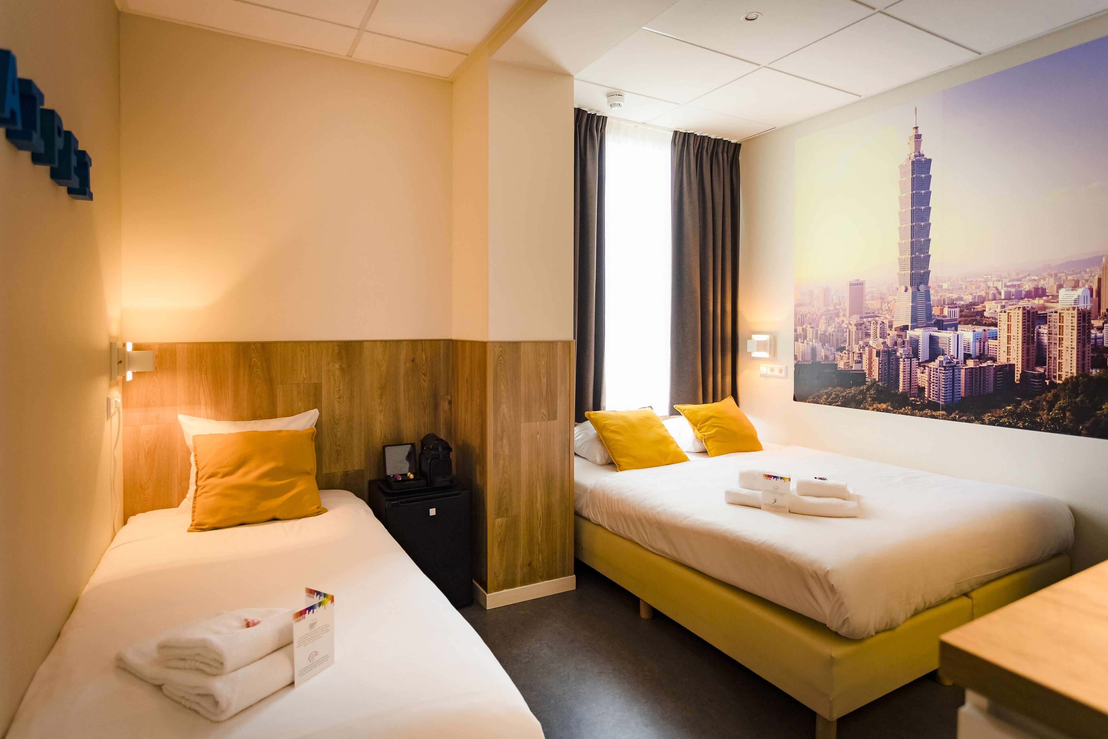 Quarto de hotel com duas camas, cabeceira de madeira, painel com um mural da paisagem urbana e uma janela com cortinas.