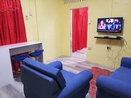 Quarto com televisão exibindo uma notícia, escrivaninha branca com duas cadeiras azuis e cortina vermelha.