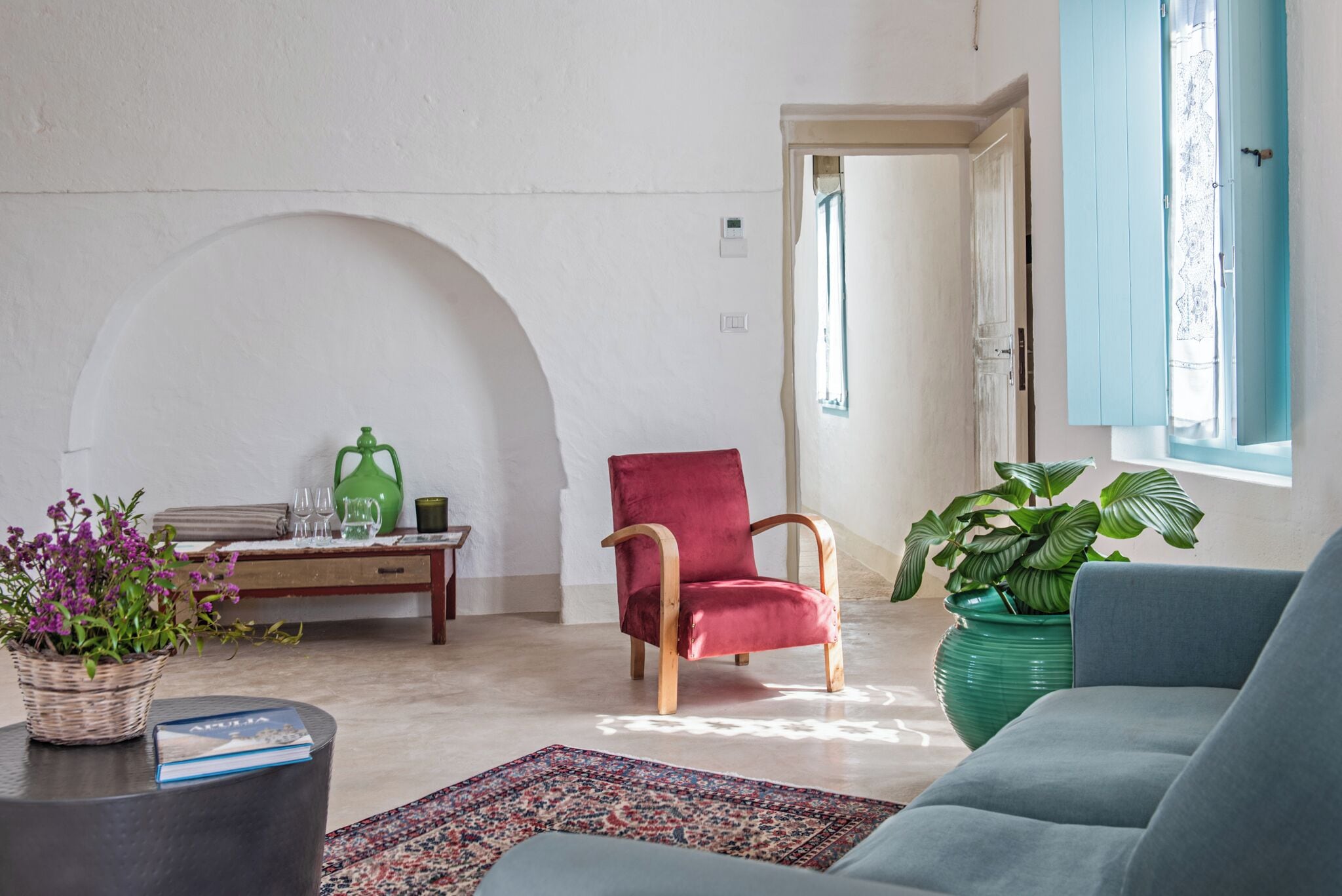 Uma sala de estar com uma cadeira vermelha, um sofá azul, uma planta verde e um tapete estampado.
