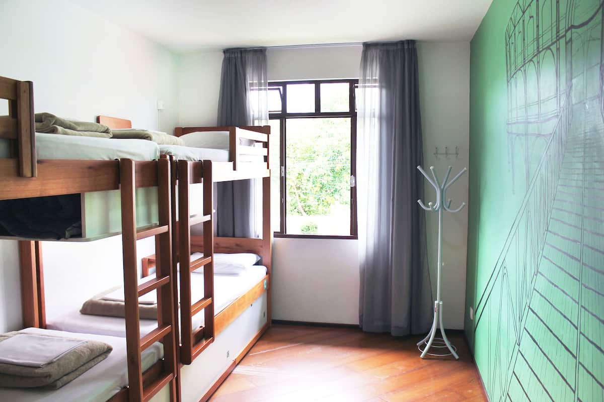 Quarto com beliches, janela com cortinas, cabideiro e parede verde com design geométrico.